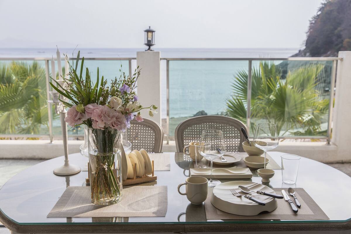 Mesa posta para dois com vista para o oceano, um vaso de flores e uma vela em uma varanda com vista para o mar.