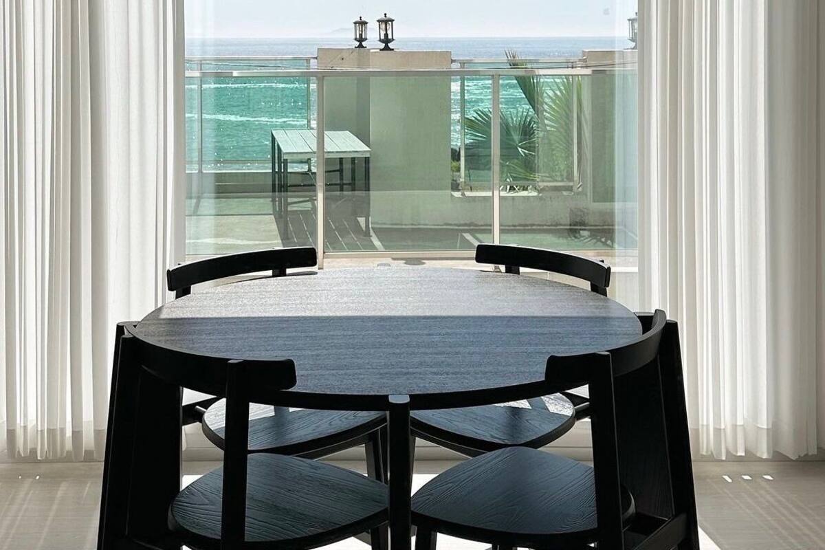 Uma sala de jantar com mesa redonda e cadeiras, janelas amplas com cortinas brancas e vista para o oceano.
