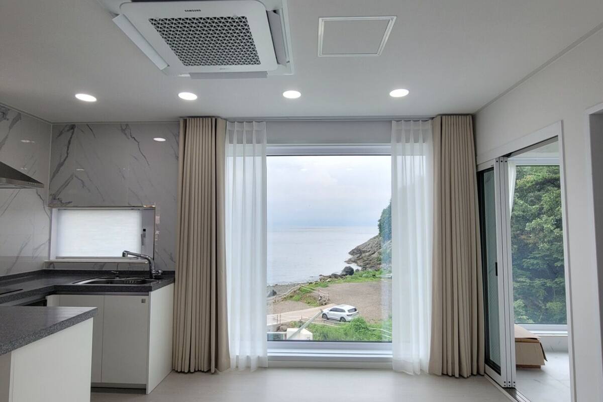 Uma cozinha moderna com fogão embutido, uma janela com cortinas e vista para o mar.