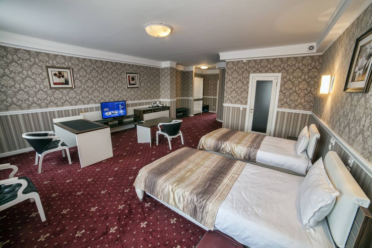 Quarto de hotel com duas camas, uma escrivaninha com computador, uma cadeira e piso coberto por um tapete vermelho.