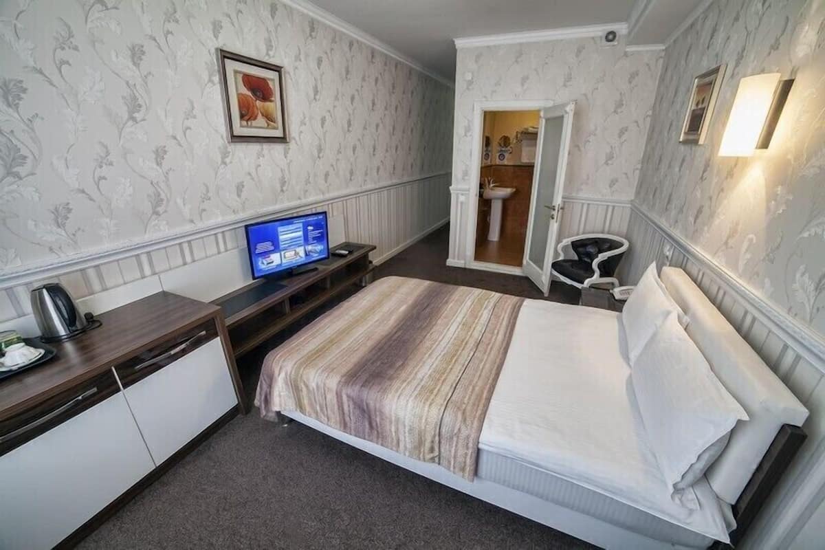 Quarto de hotel com cama, escrivaninha com computador, cadeira, mesinha com chaleira e banheiro visível por uma porta aberta.