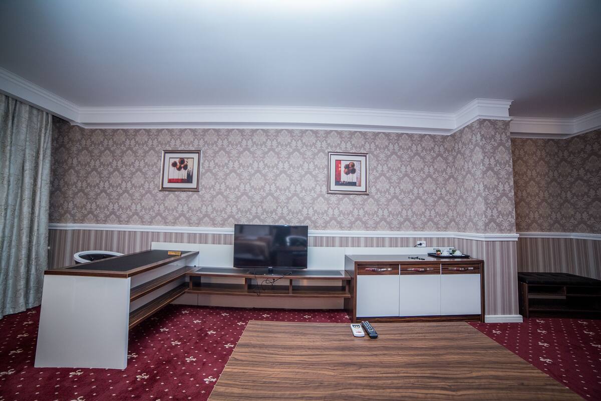 Quarto de hotel com mesa de centro de madeira, televisão em móvel, pequeno armário e carpete estampado.