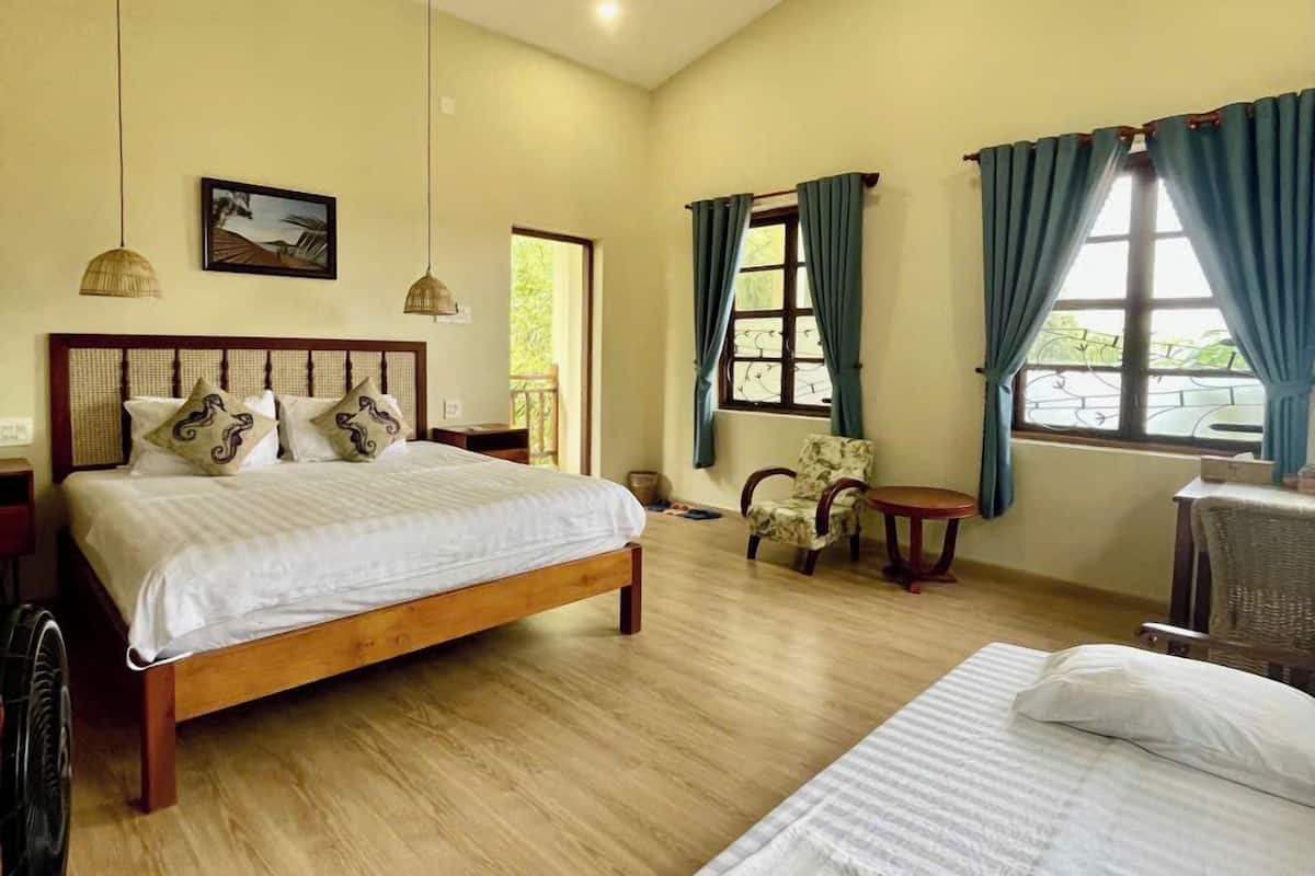 Quarto de hotel com uma cama grande, uma cama menor, um ventilador de teto, uma janela com cortinas, uma cadeira, uma mesa e um quadro na parede.