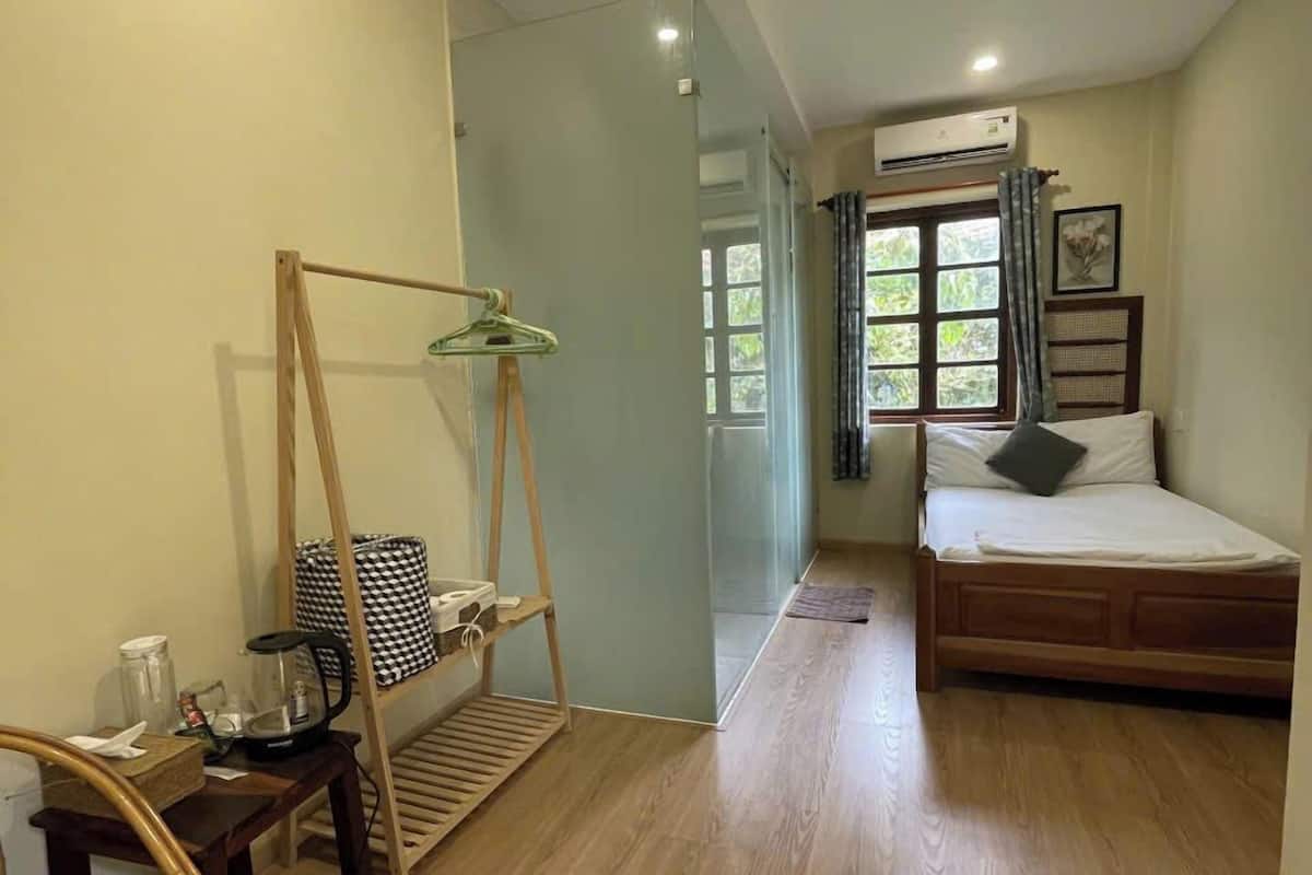 Quarto com cama de madeira, uma mesinha com abajur, uma cadeira e um cabideiro com roupas.