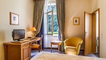 Quarto de hotel com cama, escrivaninha com computador, cadeira, mesa pequena e cortinas.
