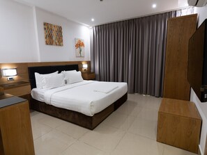 Quarto de hotel moderno com uma cama grande, cabeceira de madeira, duas mesas de cabeceira com abajures, uma janela ampla com cortinas cinzas e uma TV de tela plana fixada na parede.