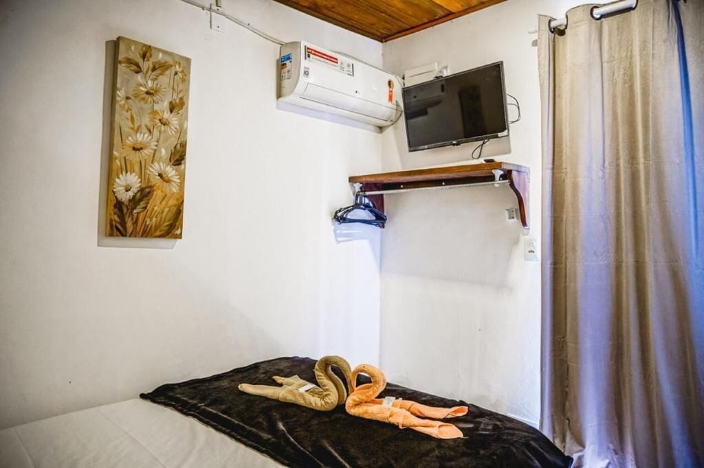 Um quarto pequeno com cama, televisão na parede, ar condicionado e um quadro.