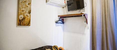 Um quarto pequeno com cama, televisão na parede, ar condicionado e um quadro.