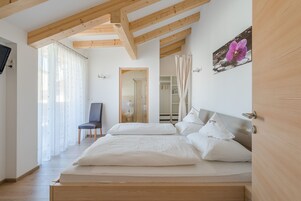 Quarto com teto de madeira, cama com roupa de cama branca, uma cadeira e um banheiro visível por uma porta aberta.