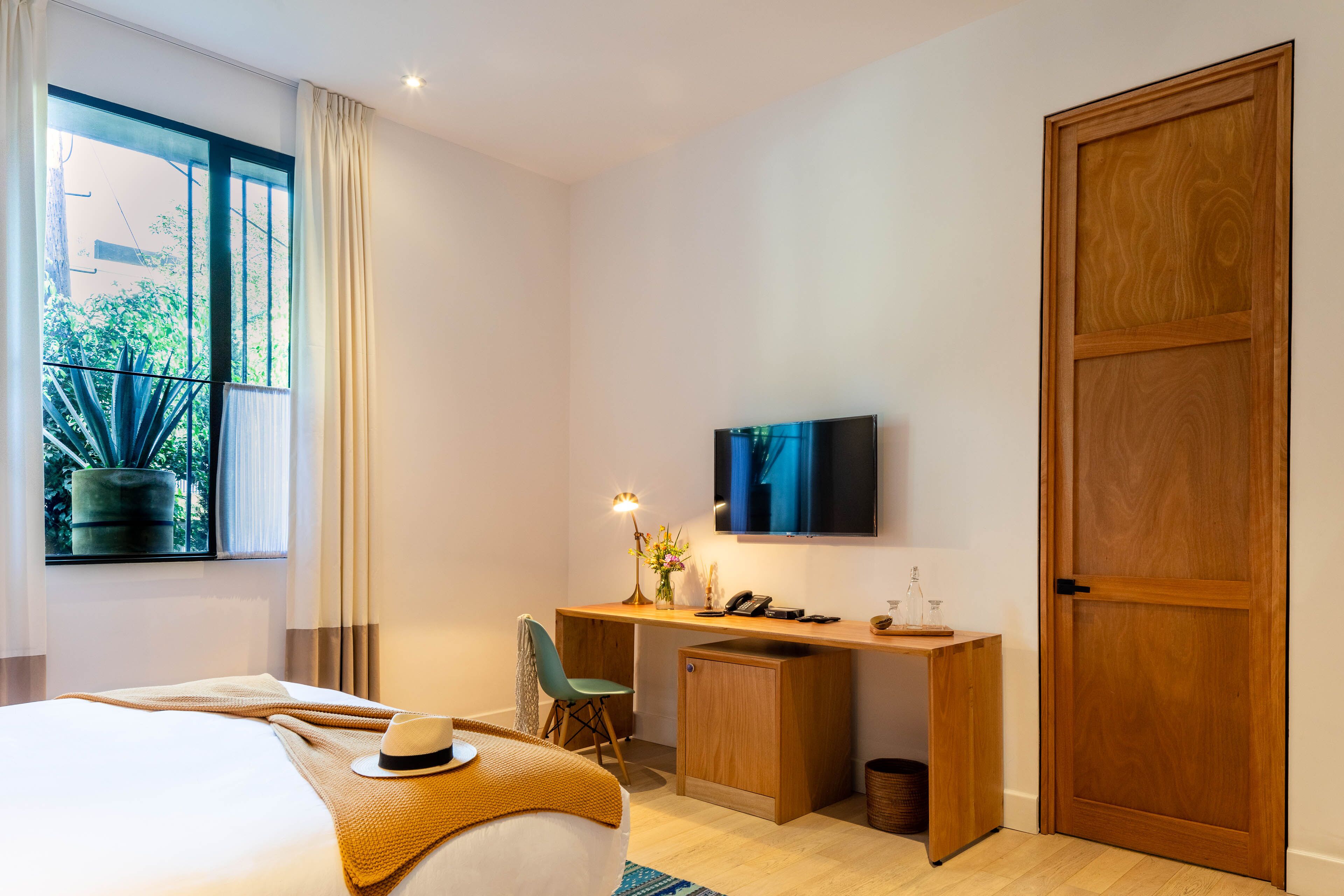 Quarto de hotel com cama, escrivaninha com televisão, janela com cortinas e porta de madeira.