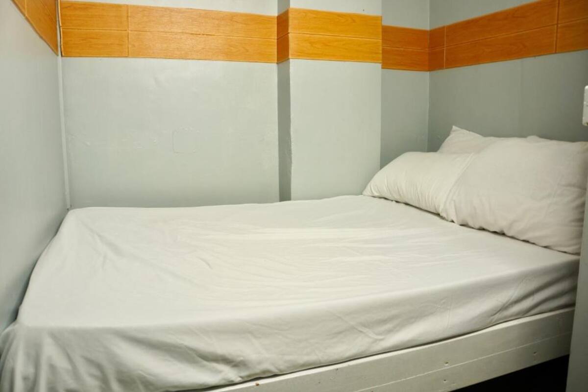 Quarto individual com uma cama de solteiro, cabeceira de madeira, roupa de cama branca e parede lisa.