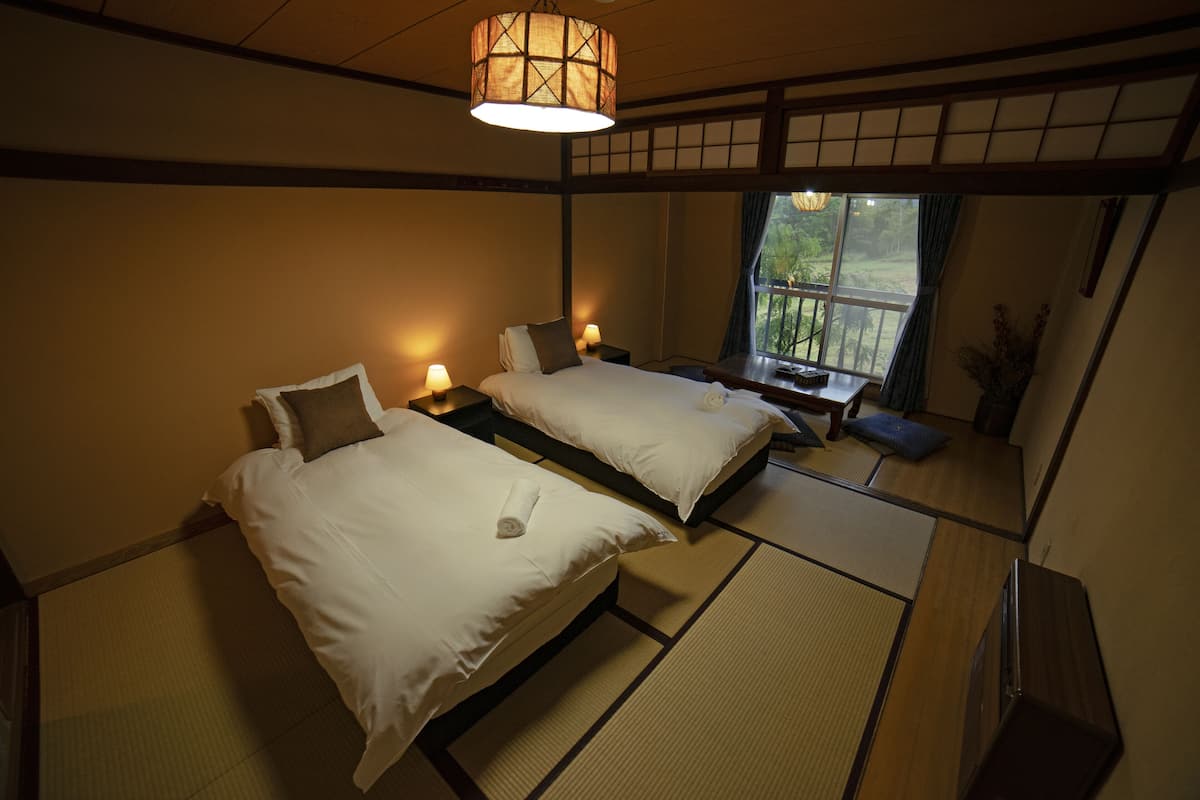 Um quarto tradicional japonês com tatames, duas camas com roupa de cama branca, um lampião pendurado e vista para o verde pela janela.