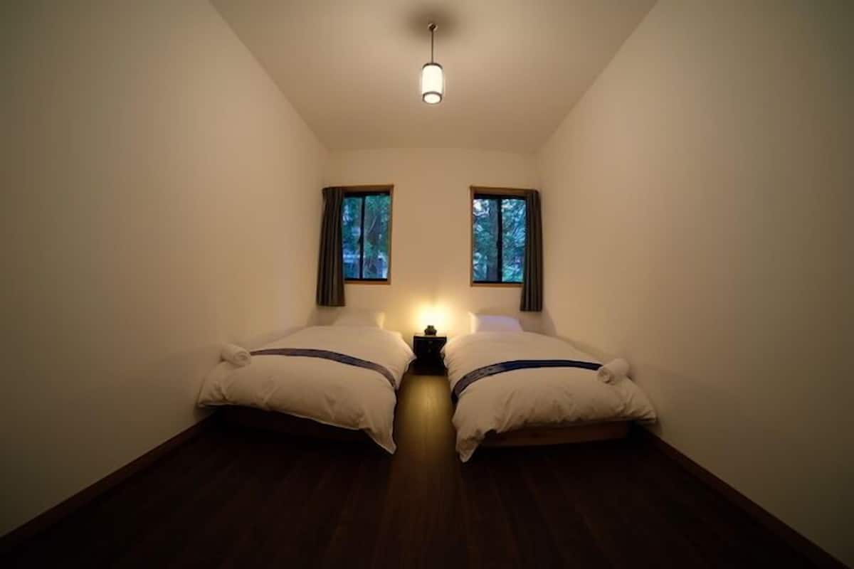 Quarto pequeno, com pouca luz, com duas camas, uma mesa de cabeceira e duas janelas com cortinas.