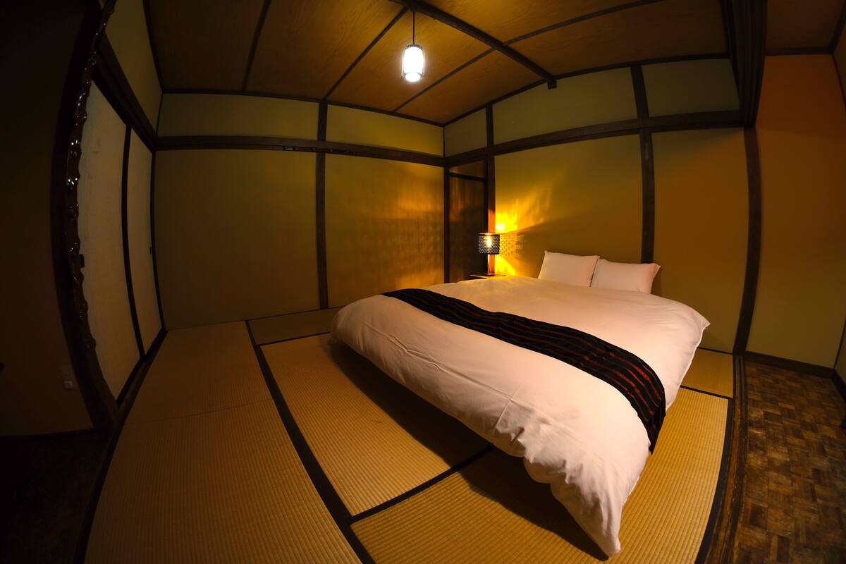 Um quarto tradicional japonês com tatames, uma cama com roupa de cama branca e uma luminária fixada na parede.