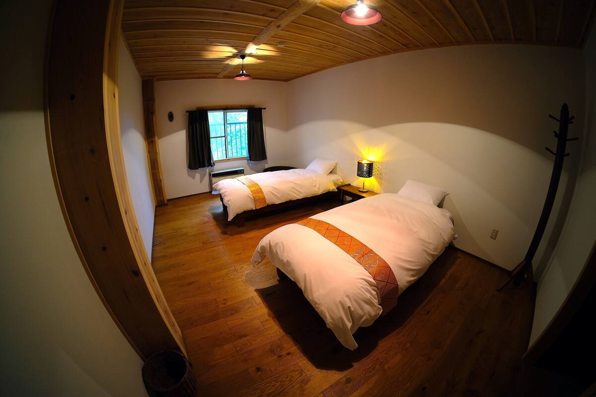Quarto com duas camas, piso de madeira, janela com cortinas e uma luminária do lado direito.
