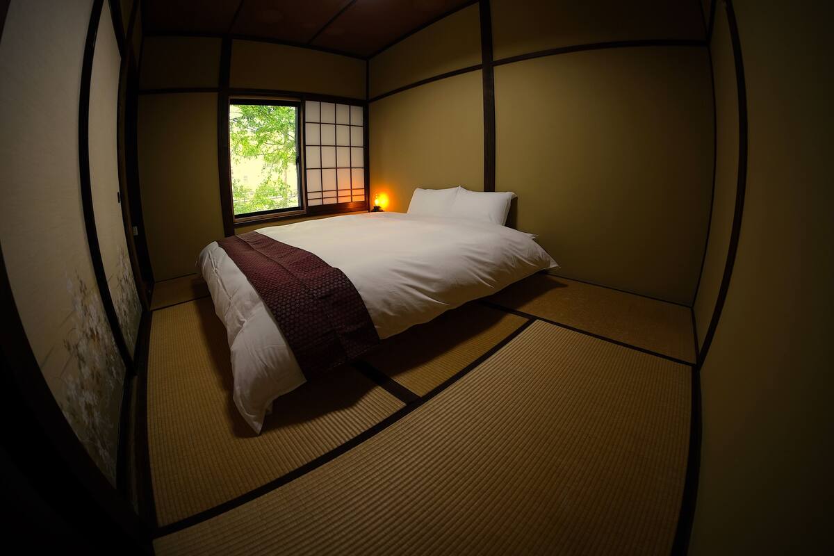 Quarto japonês tradicional com tatames, cama com roupa de cama branca e janela com vista para vegetação.