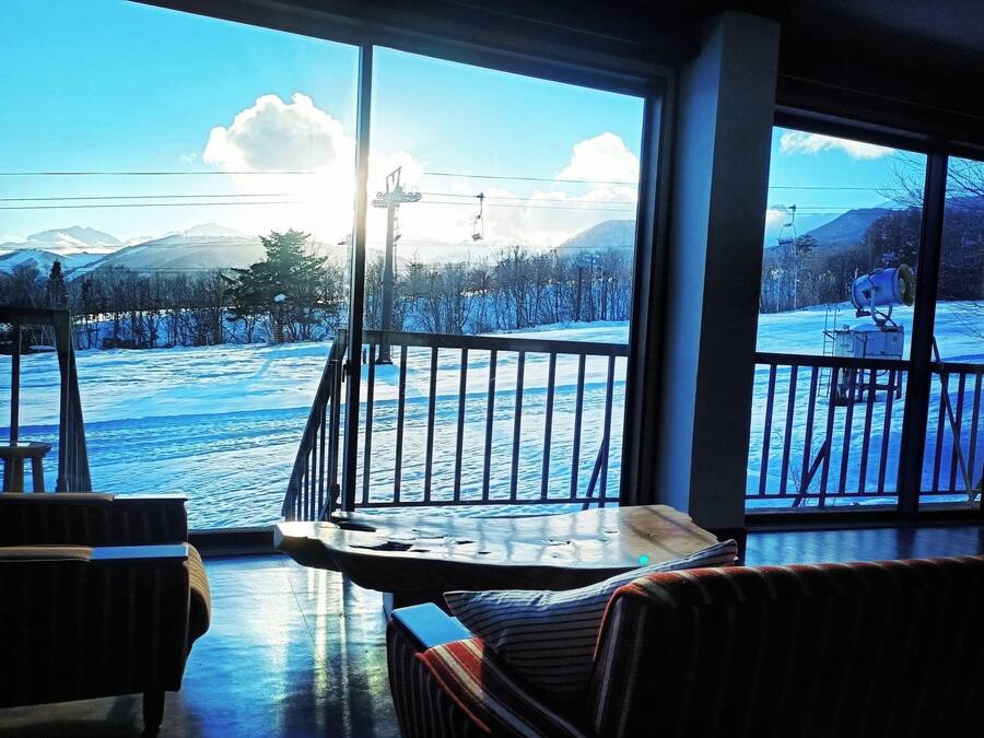 Quarto com uma grande janela com vista para uma paisagem nevada, varanda com corrimão e área de estar com mesa e cadeiras.