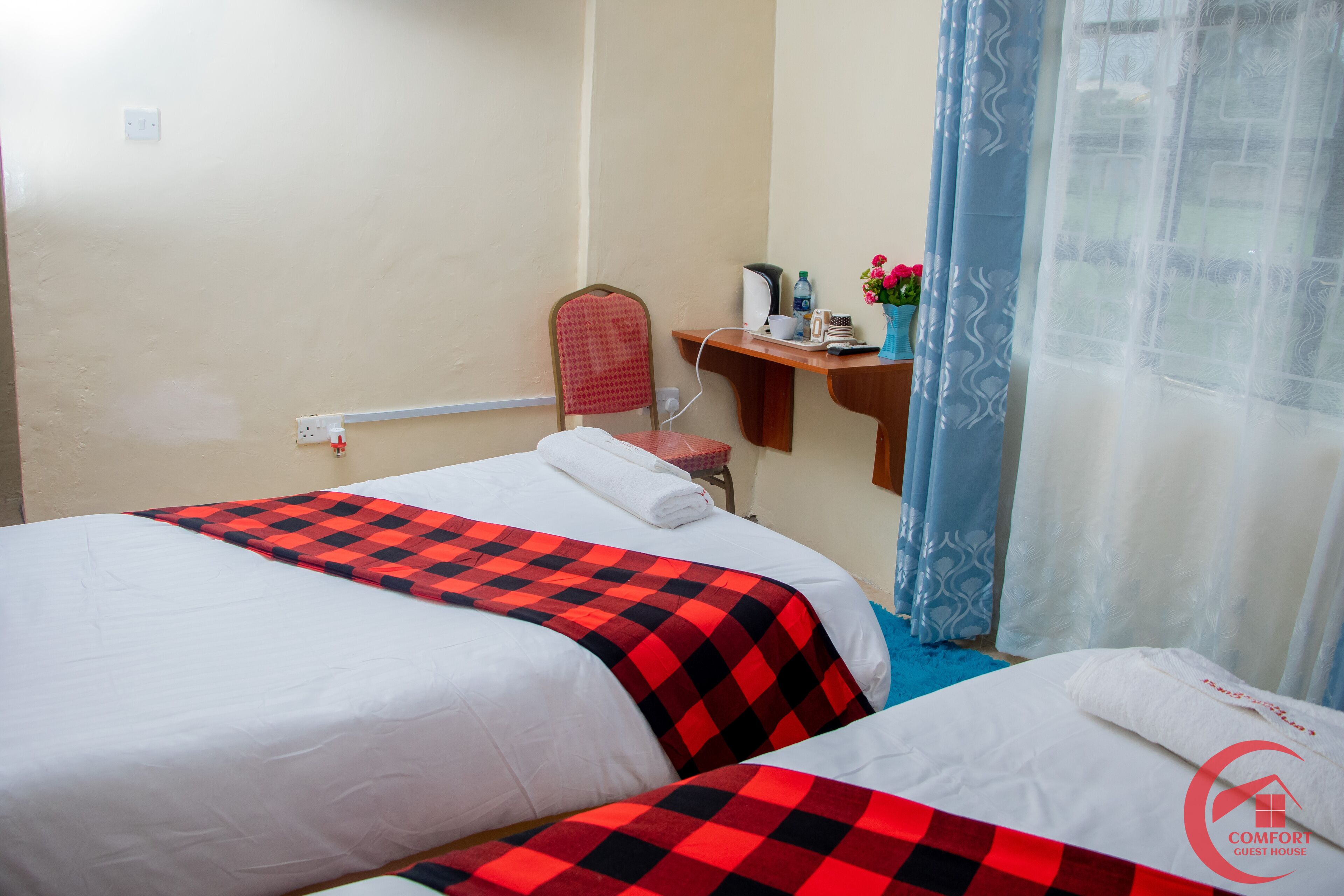 Quarto de hotel com duas camas, uma pequena escrivaninha com cadeira, janela com cortinas e uma tomada na parede.