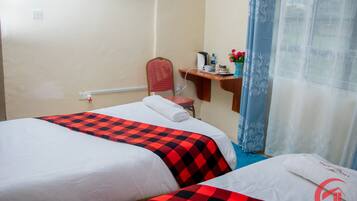 Quarto de hotel com duas camas, uma pequena escrivaninha com cadeira, janela com cortinas e uma tomada na parede.