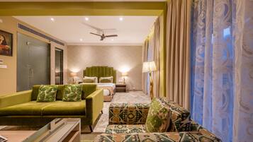Quarto de hotel com um sofá verde, um banquinho estampado, uma cama com cabeceira verde, um ventilador de teto e uma grande janela com cortinas.