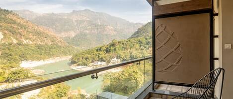 Uma varanda com vista para um rio e montanhas, uma mesa com cadeiras e uma porta decorativa.