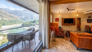 Quarto com varanda e vista para um rio e montanhas, mobiliado com cadeiras de vime e um sofá laranja.