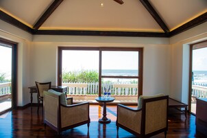 Quarto com uma grande janela com vista para o oceano, mobiliado com cadeiras de vime e uma pequena mesa.
