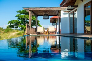 Piscina com deck de madeira e área de estar ao ar livre, cercada por um edifício branco com grandes janelas.