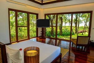 Quarto com uma cama grande, televisão e vista para um jardim tropical através de janelas amplas.