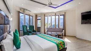 Quarto de hotel com uma cama, duas cadeiras, um sofá, uma TV e amplas janelas com vista para as montanhas.