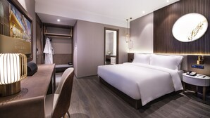 Quarto de hotel moderno com uma cama grande, uma escrivaninha com luminária, uma cadeira, um espelho e um ponto de luz fixado na parede.