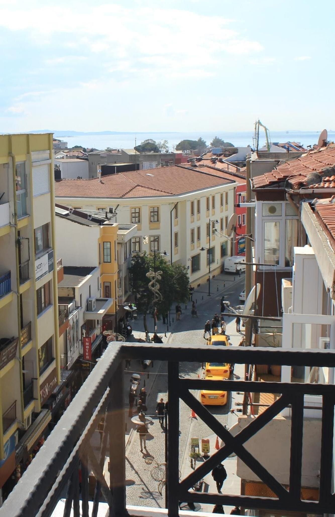 Vista de um ponto alto sobre uma rua da cidade, com edifícios, uma praça central e um corpo de água ao longe.