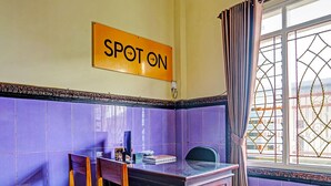 Quarto com paredes de azulejos roxos, uma escrivaninha de madeira com cadeira, uma janela com cortinas e um letreiro com a inscrição 'SPOT ON'.