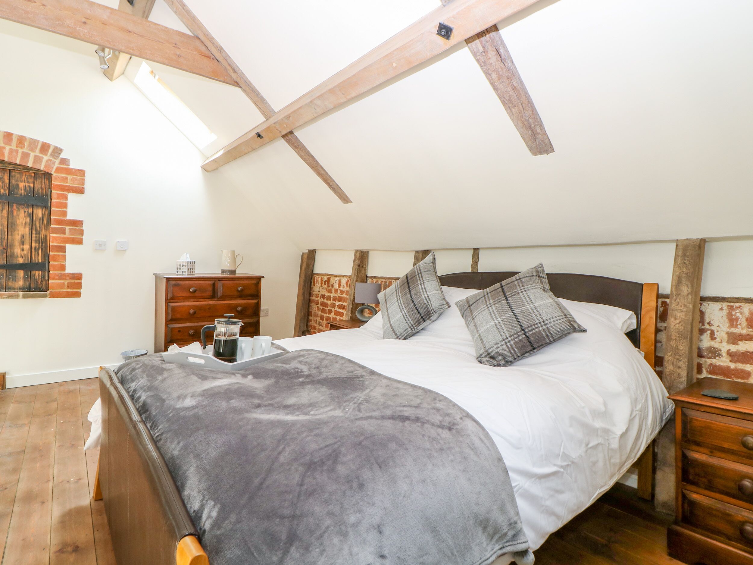 Quarto com teto inclinado, vigas de madeira aparente, parede de tijolo, cômoda de madeira, mesa de cabeceira e cama com manta cinza e almofadas xadrez.