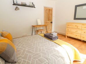 Quarto com cama, cômoda de madeira, mesa pequena e prateleira com livros e um vaso.