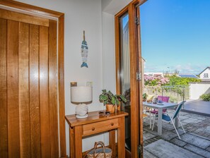 Uma porta de madeira com uma pequena mesa ao lado, uma luminária fixada na parede e vista para um jardim com flores cor-de-rosa e um painel solar.