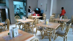 Restaurante com mesas e cadeiras de madeira, área de cozinha e uma pessoa vestindo jaqueta vermelha ao fundo.
