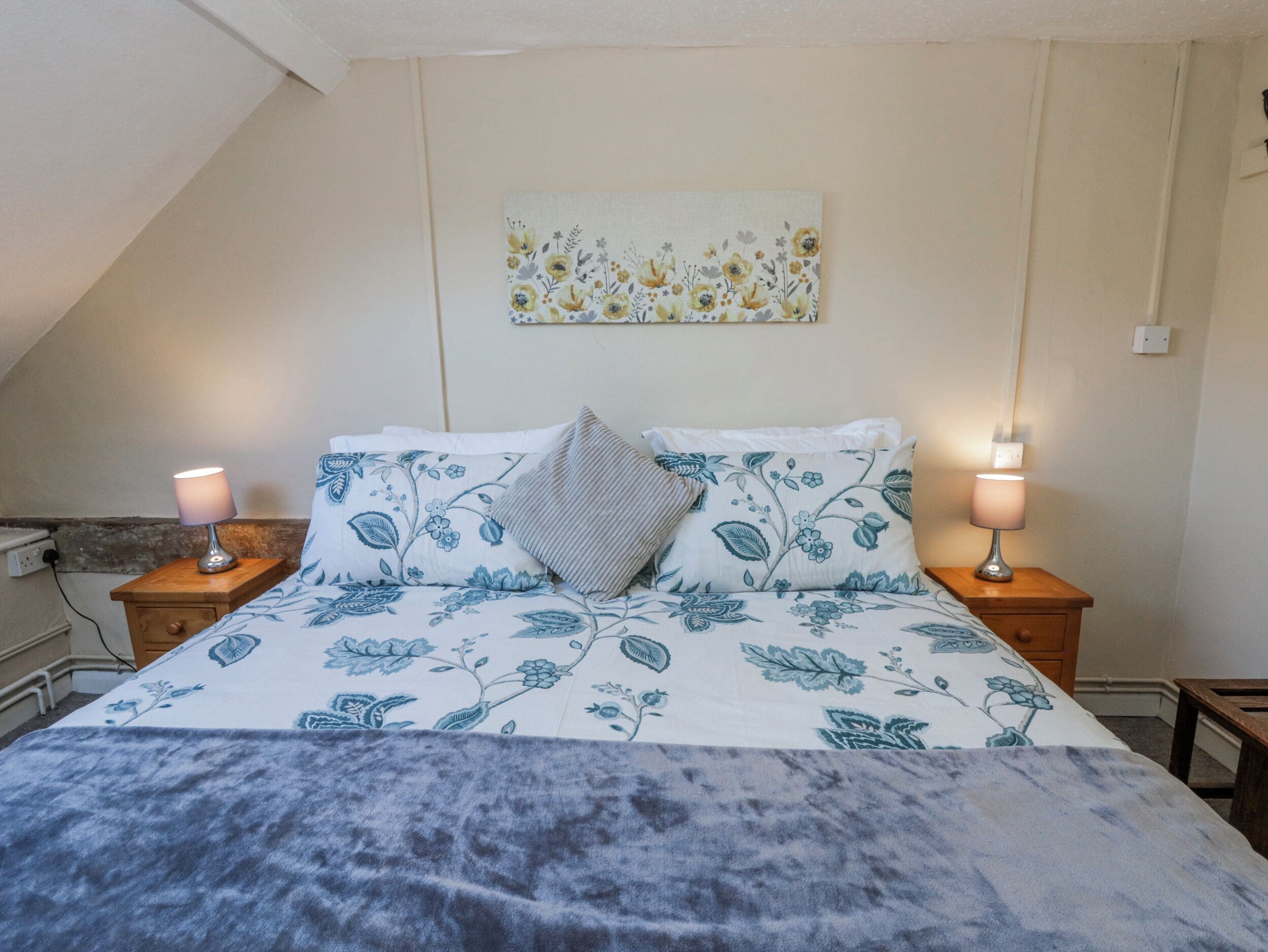 Quarto com cama, duas luminárias de cabeceira, uma pintura floral na parede e uma mesinha de cabeceira de madeira.