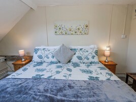 Quarto com cama, duas luminárias de cabeceira, uma pintura floral na parede e uma mesinha de cabeceira de madeira.