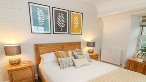 Quarto com cama de madeira, duas mesinhas de cabeceira, um radiador e três quadros com folhas na parede.