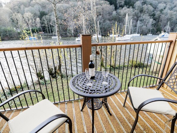 Varanda com mesa e duas cadeiras, com vista para um lago com barcos e árvores.