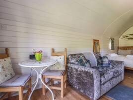 Um quarto aconchegante com piso de madeira, uma mesa redonda com cadeiras, um sofá com almofadas estampadas e uma cama com travesseiros.