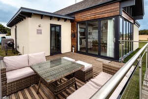 Uma área de estar externa moderna, com móveis de vime, uma mesa de vidro e vista para o interior através de grandes portas deslizantes.