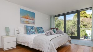 Quarto com cama, mesa de cabeceira, pintura e vista para o verde através de uma grande janela.