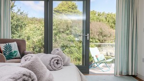 Quarto com cama, toalhas dobradas, uma cadeira e vista para o verde através de uma porta de correr de vidro.