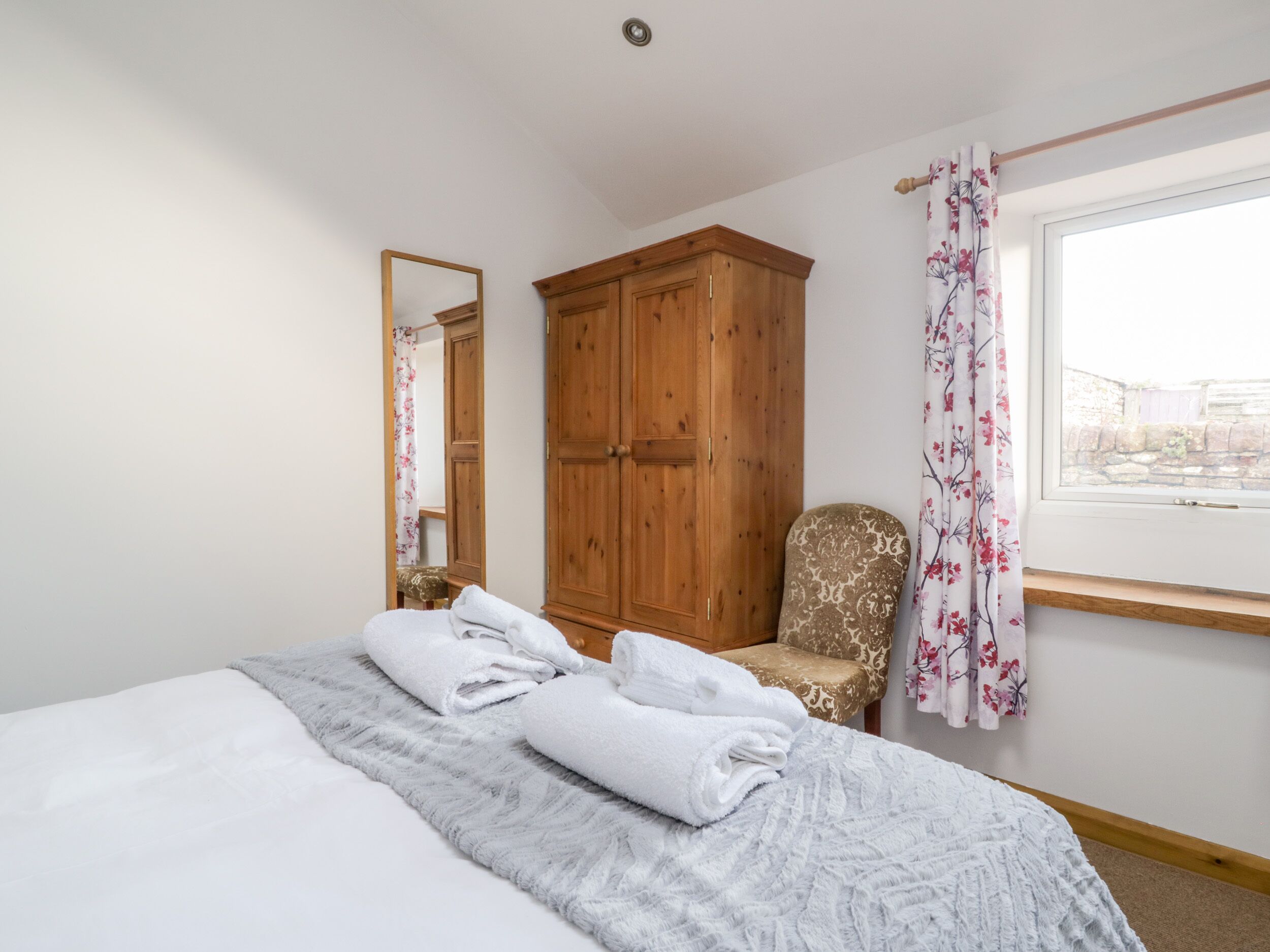 Um quarto com cama, guarda-roupa de madeira, cadeira, janela com cortinas e espelho.