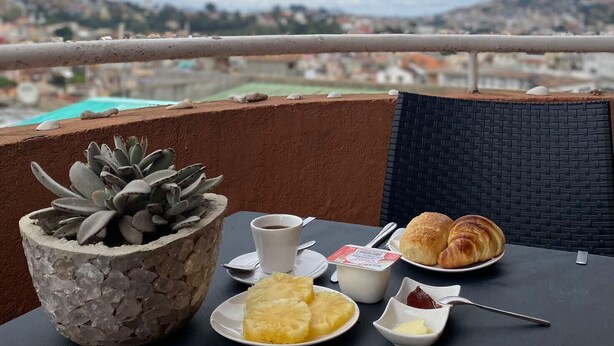 Mesa com xícara de café, croissants, fatias de abacaxi e manteiga numa varanda com vista para a paisagem urbana.