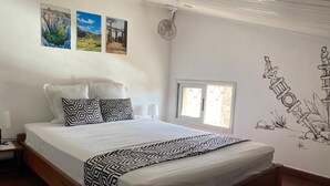 Quarto com cama de madeira, roupa de cama branca, almofadas estampadas, ventoinha na parede e três quadros de paisagens.
