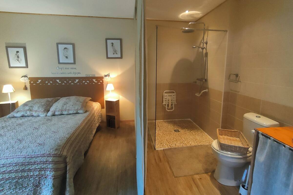 Quarto com cama, luminárias de cabeceira e obras de arte na parede. Ao lado, um banheiro com chuveiro, vaso sanitário e uma pequena cesta.