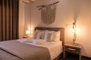 Uma cama bem arrumada com colcha estampada, uma decoração de macramé pendurada na parede e um abajur sobre uma mesinha de cabeceira de madeira.